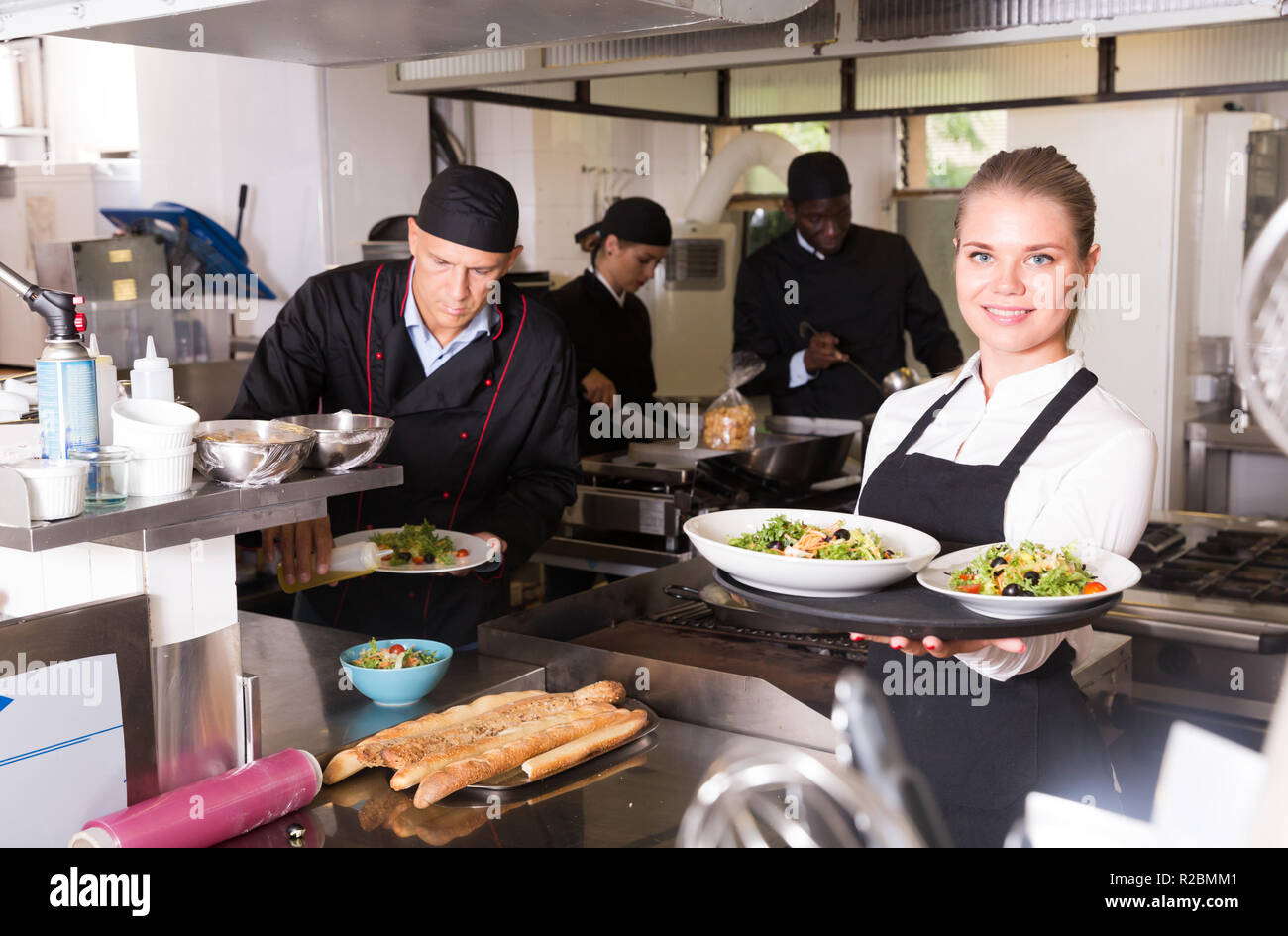 Portrait of attractive waitress with ordered dishes in kitchen of ...