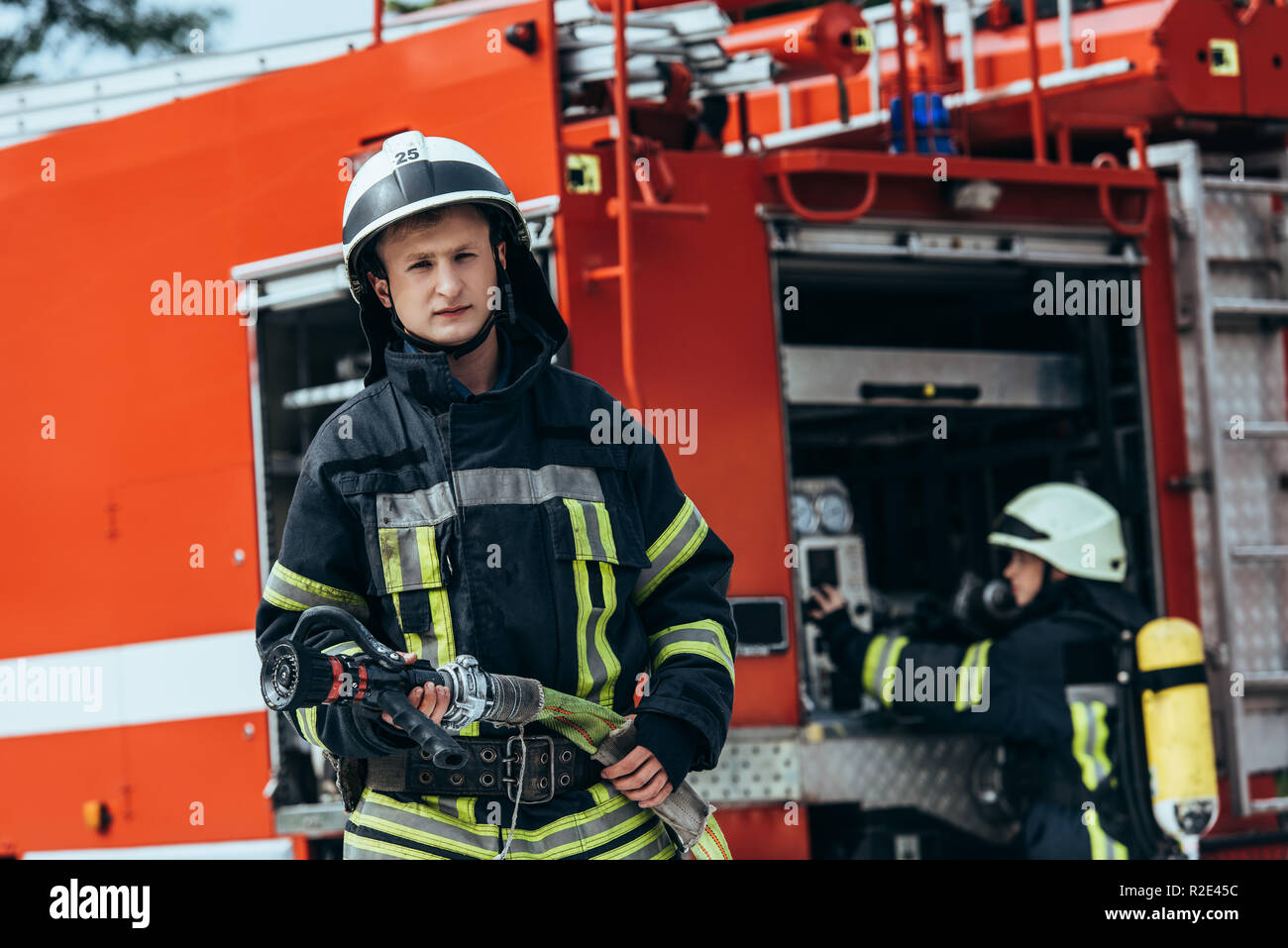 male firefighter in uniform holding water hose while colleague checking ...