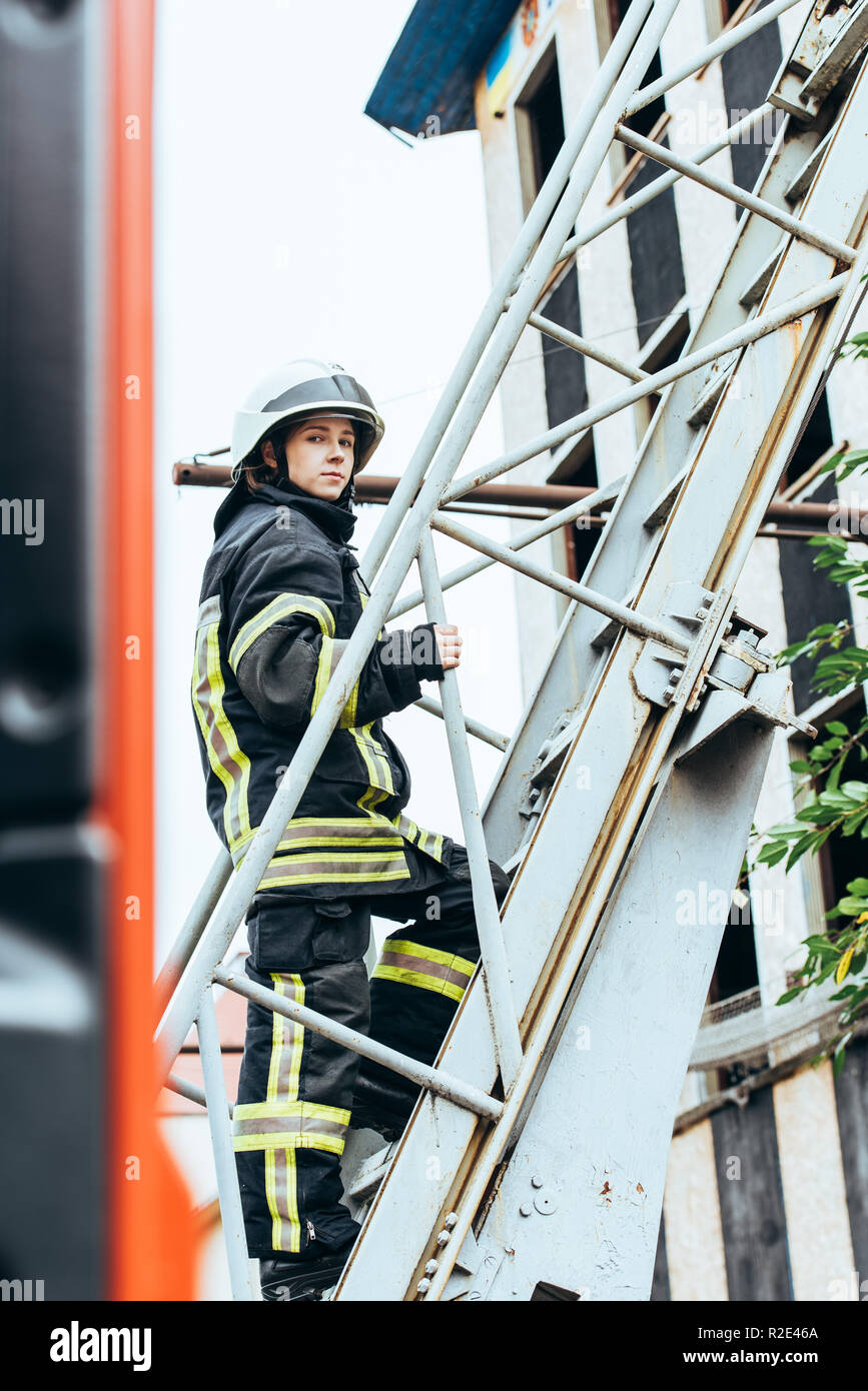 female firefighter in protective uniform and helmet standing on ladder ...