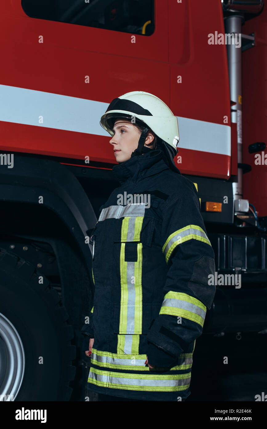 side view of female firefighter in helmet standing at fire station ...