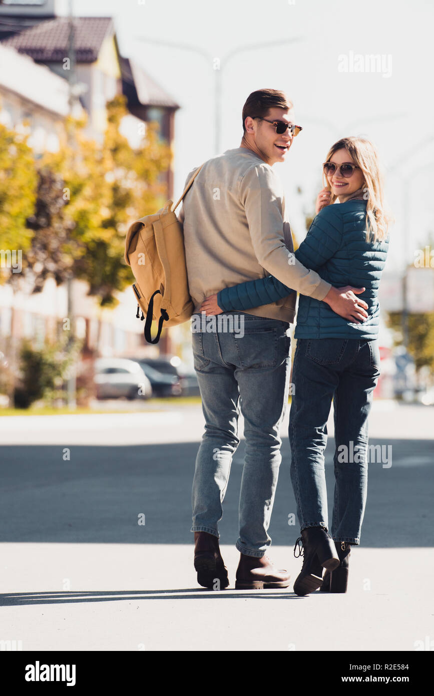 back view of couple hugging and looking at camera while walking on ...