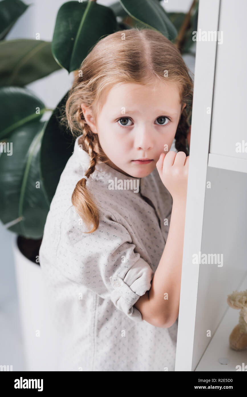 scared little child standing behind bookshelves and looking at camera ...