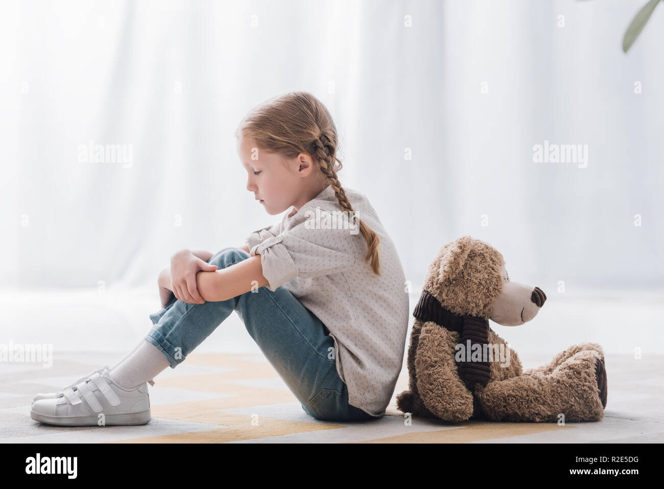 side view of sad little child sitting on floor back to back with teddy ...