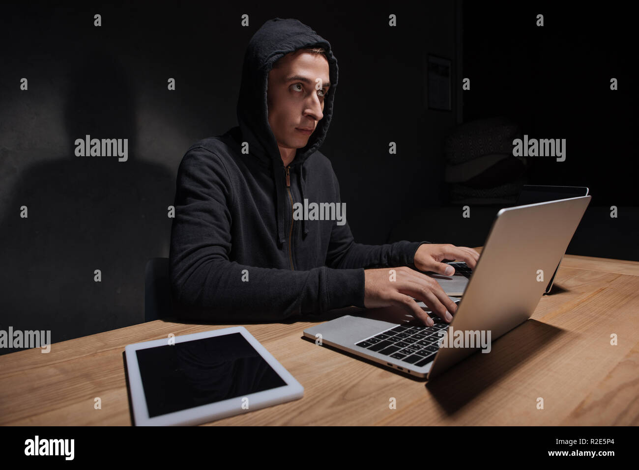 hacker in black hoodie using laptops at tabletop with tablet in dark ...