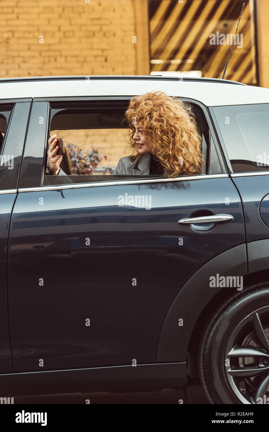side view of curly redhead woman leaning out from car and taking selfie ...