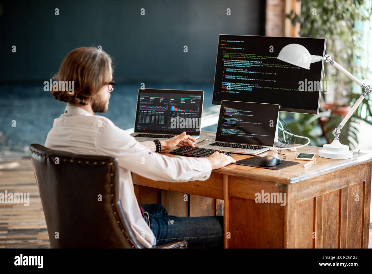 Young male programmer writing a program code sitting at the workplace with three monitors in the ...