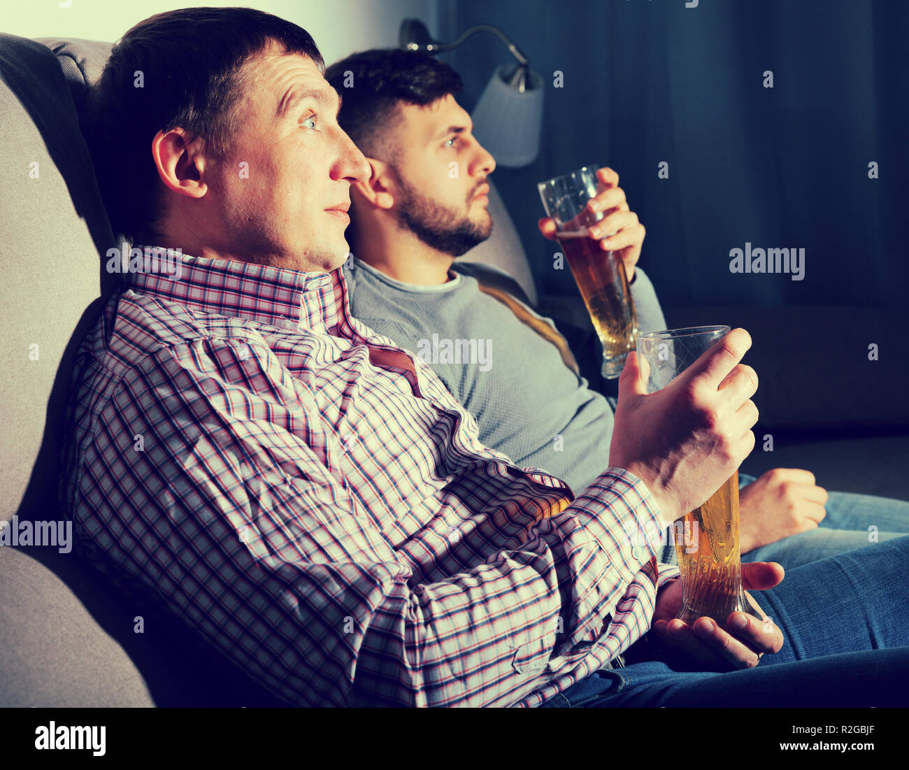 Two men resting at home, sitting on sofa with glass of beer, watching ...