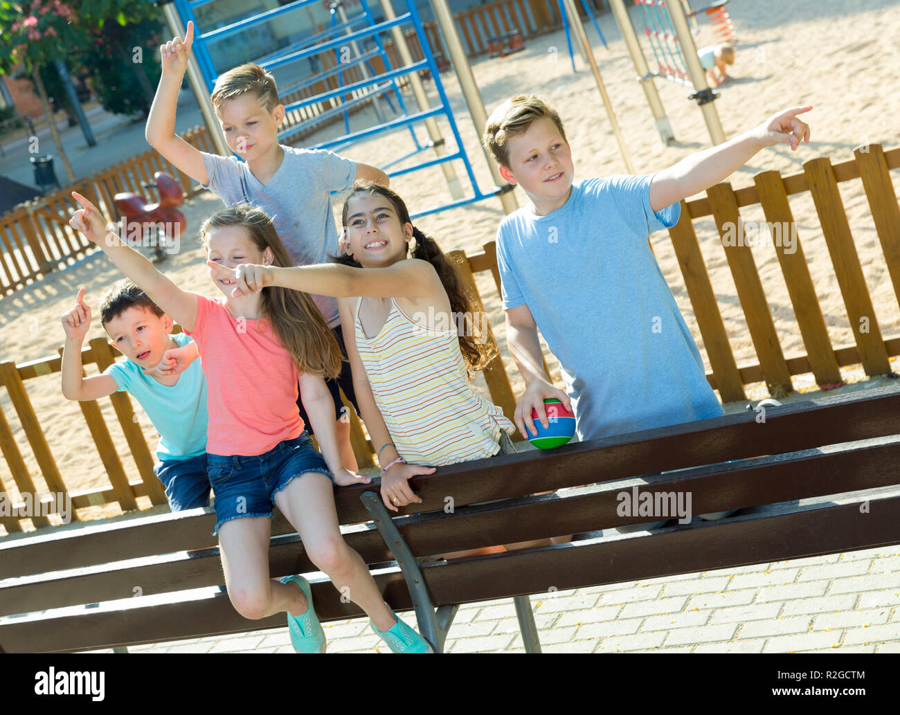 Five glad children sitting on a bench at the playground and point to ...
