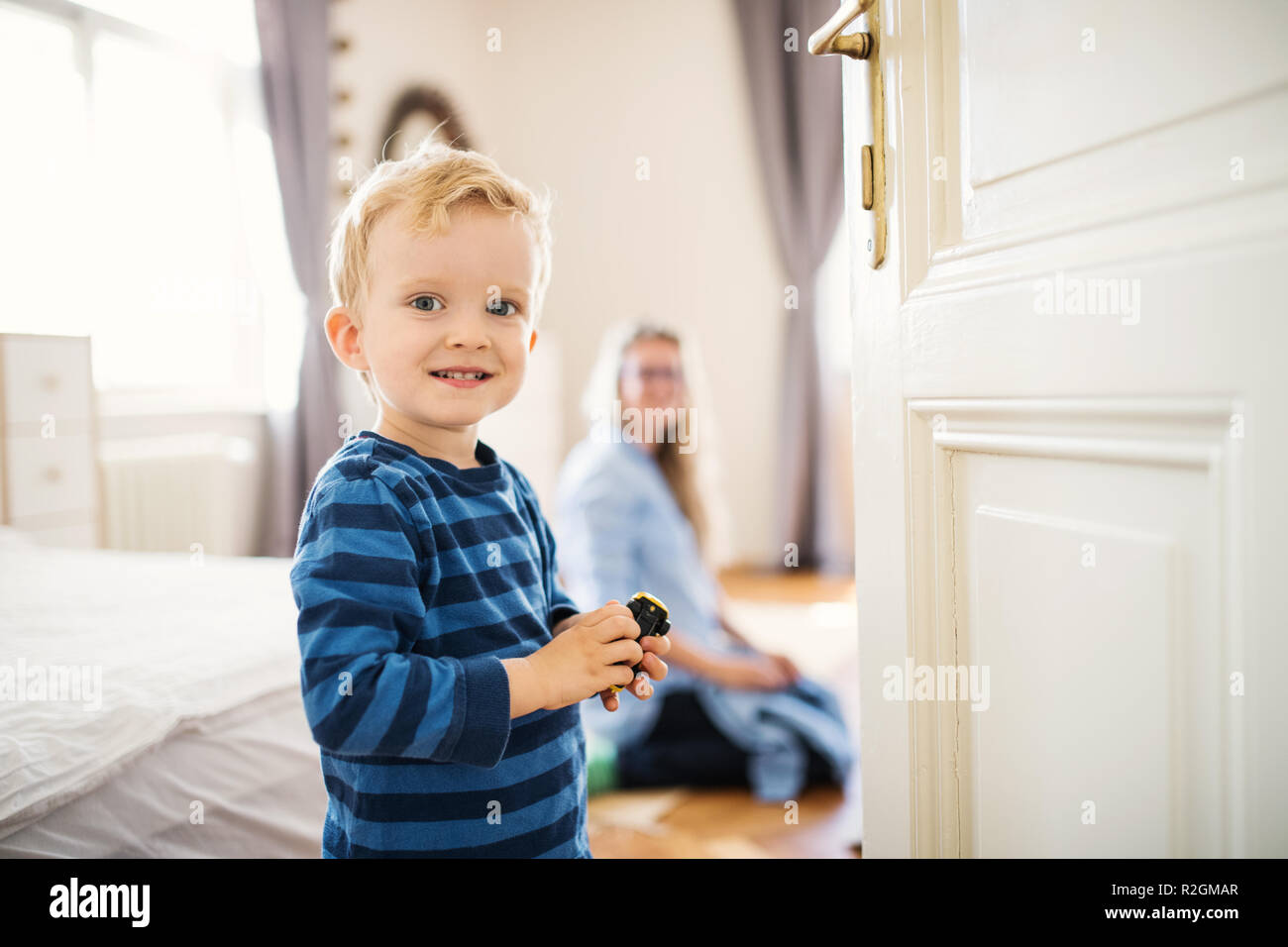 A toddler boy with young mother in the background inside in a bedroom ...