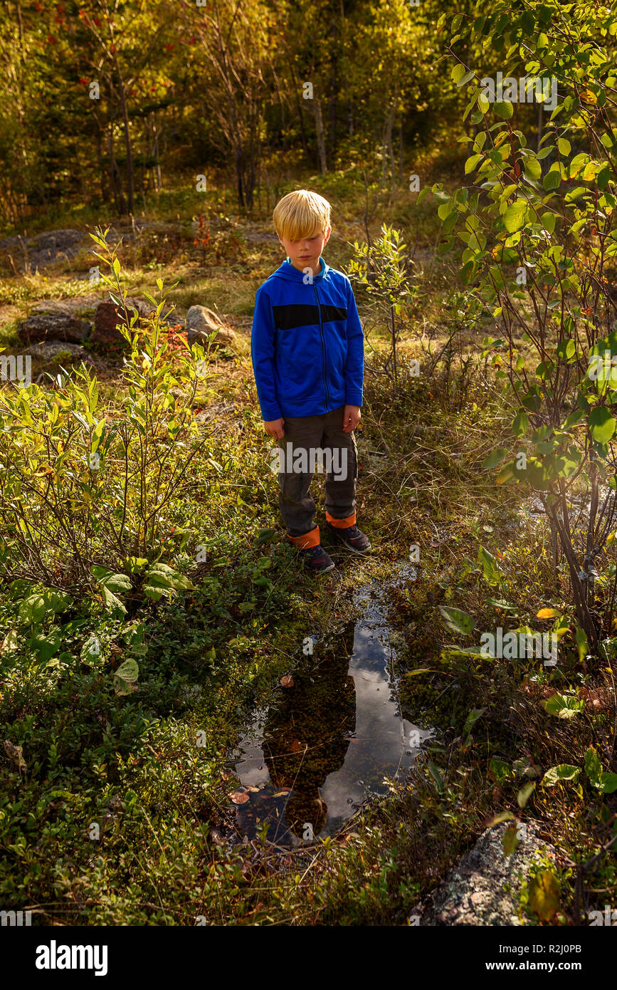 Boy standing in the forest looking at his reflection in a puddle of ...