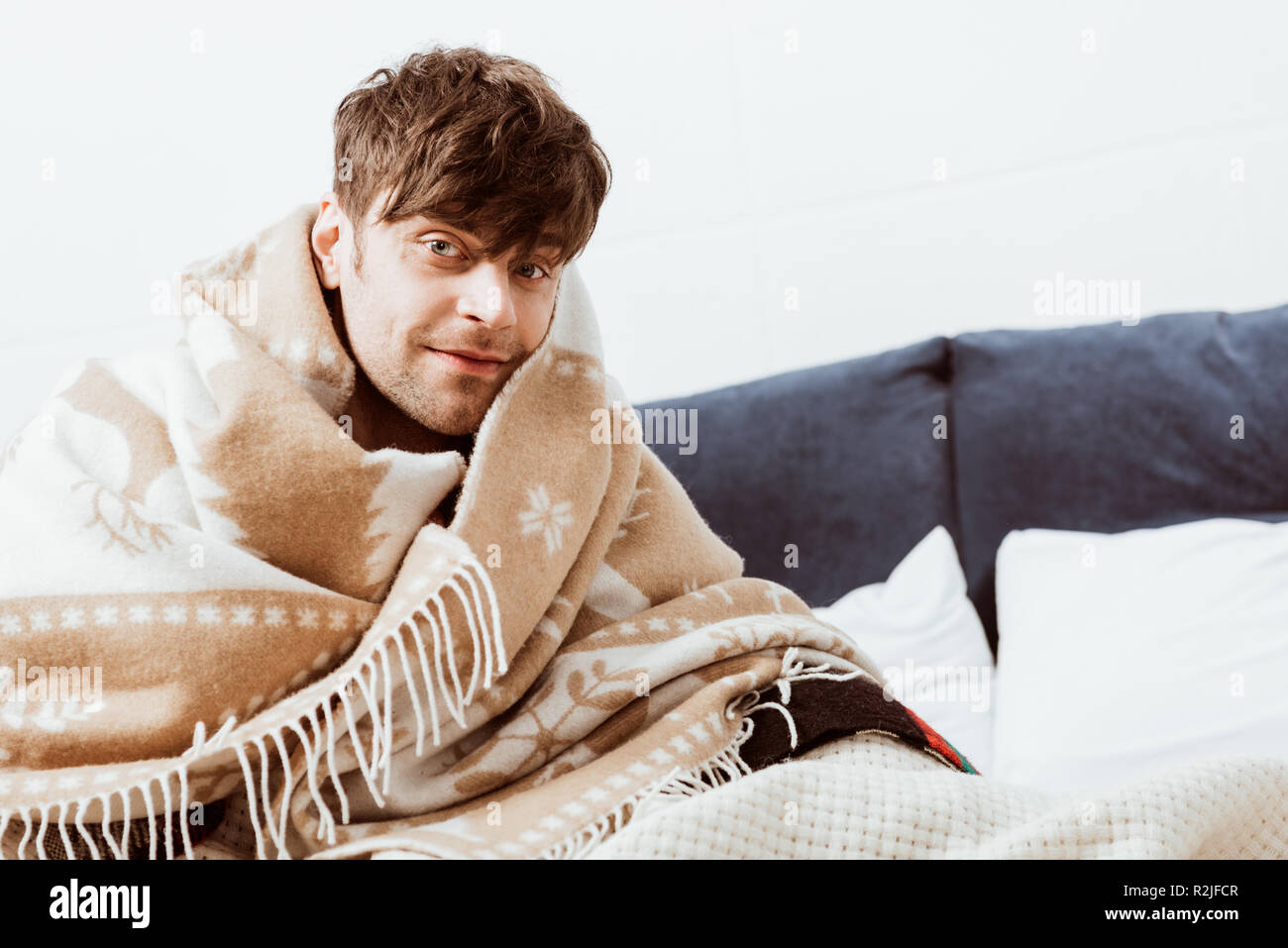 cheerful sick young man wrapped in blanket sitting on bed at home Stock ...