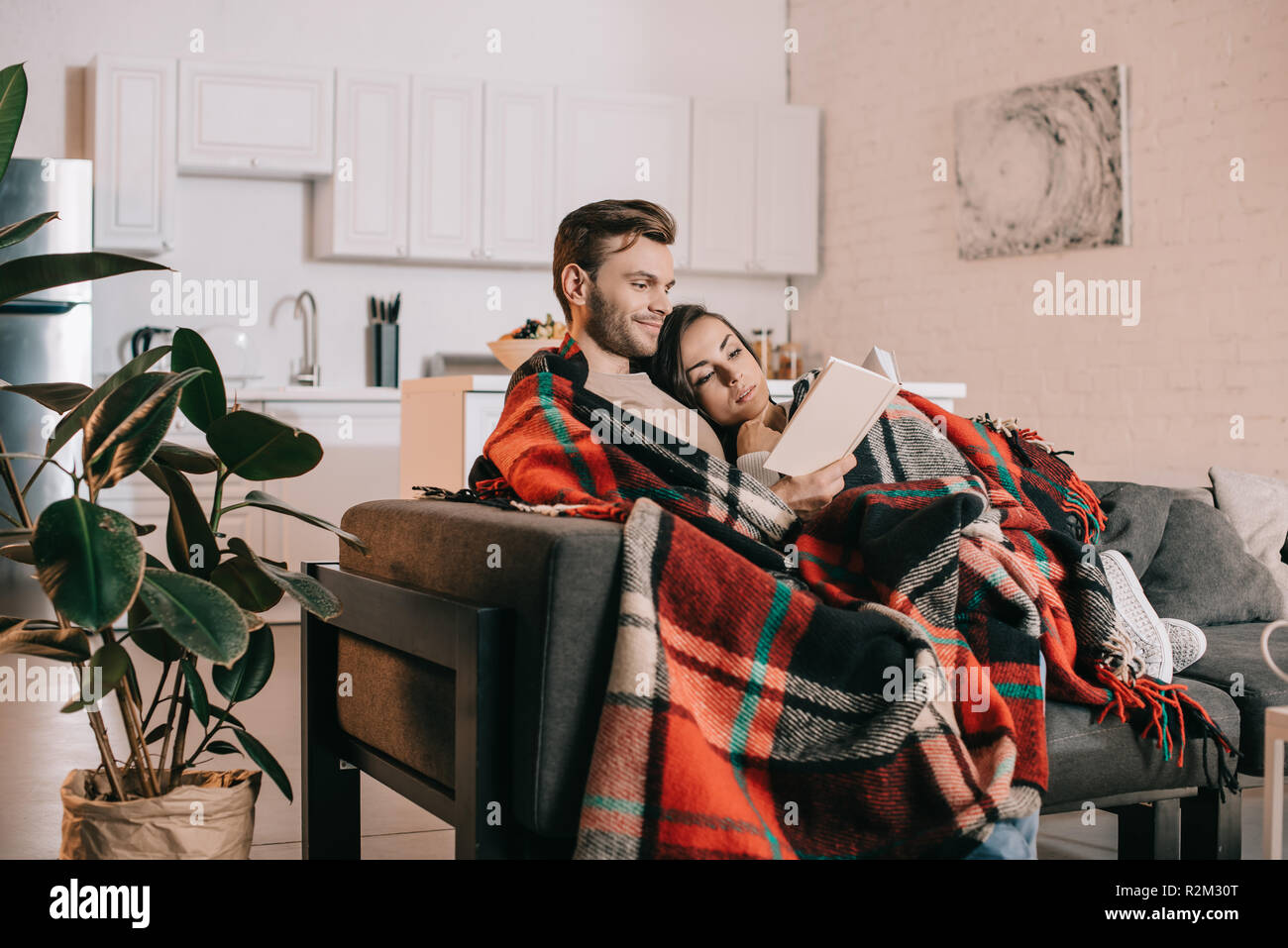 Smiling Young Couple Reading Book Together On Couch Under Plaid At Home