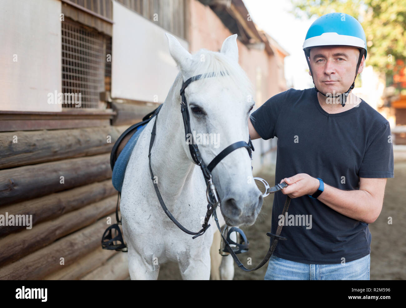 Mature smiling man farmer in helmet standing with white horse at stable ...