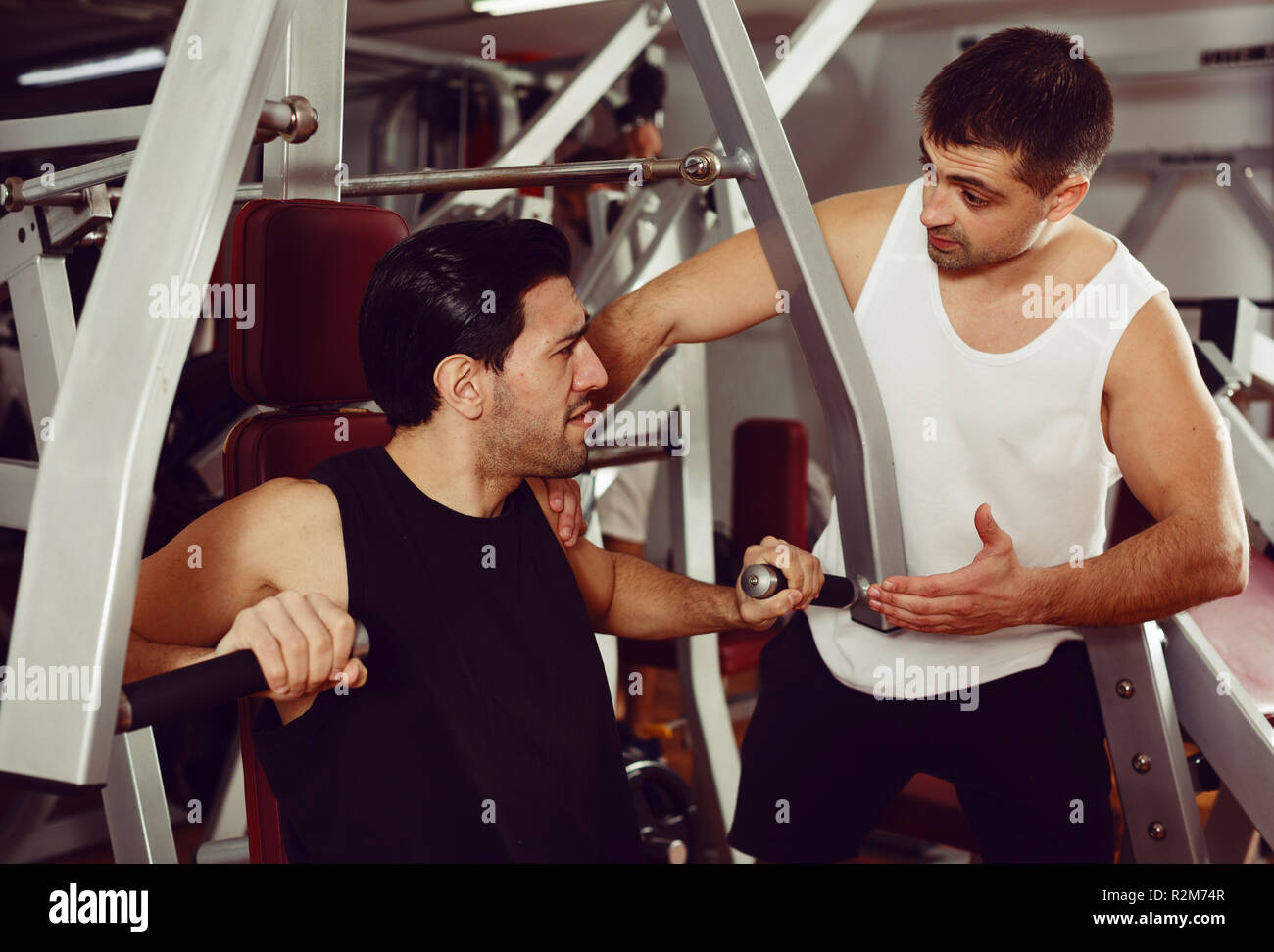 Athletic man helping friend to performing exercises on fitness machine ...