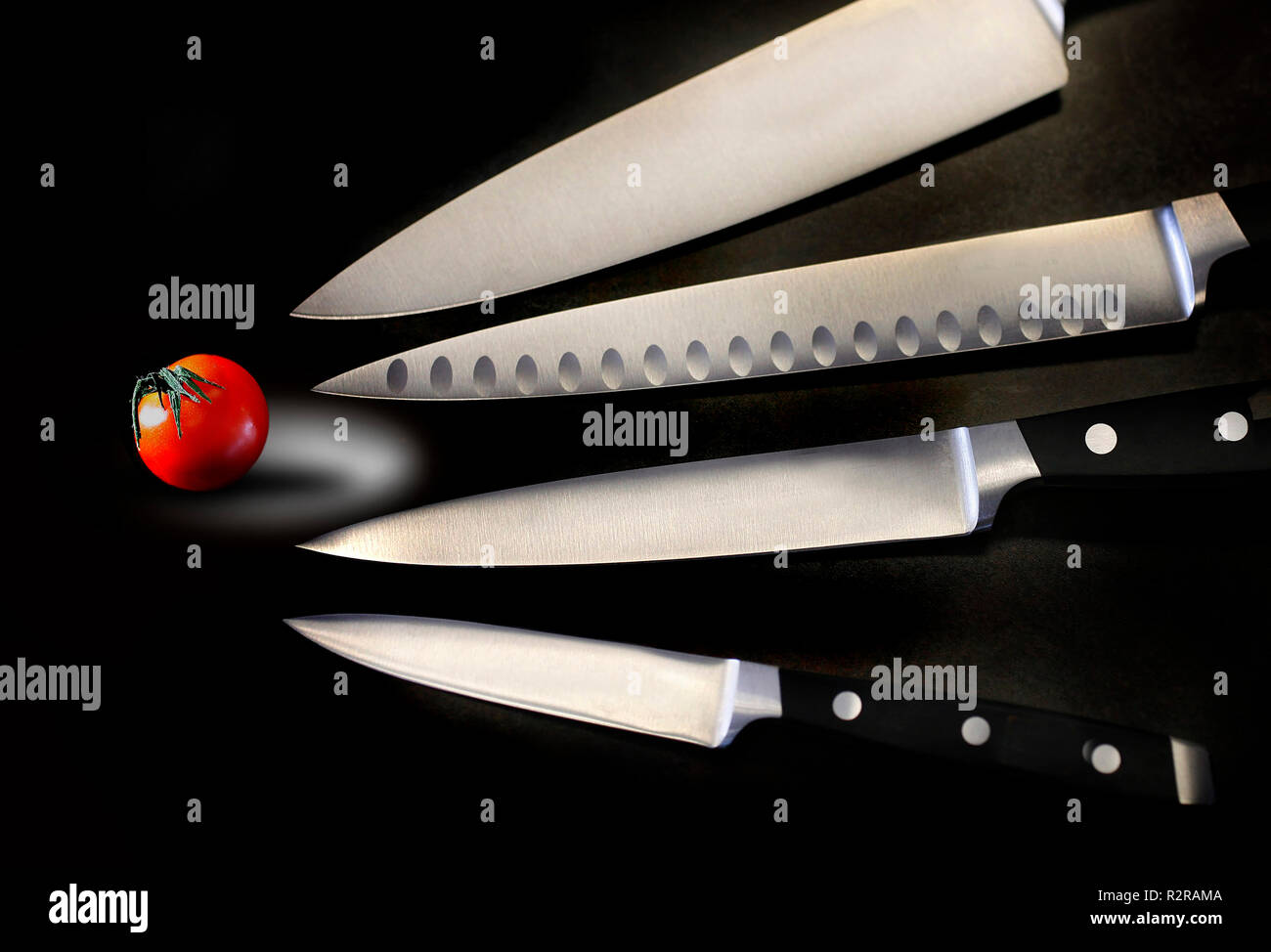 Assorted sharp and shiny kitchen knives are seen with a small tomato in ...