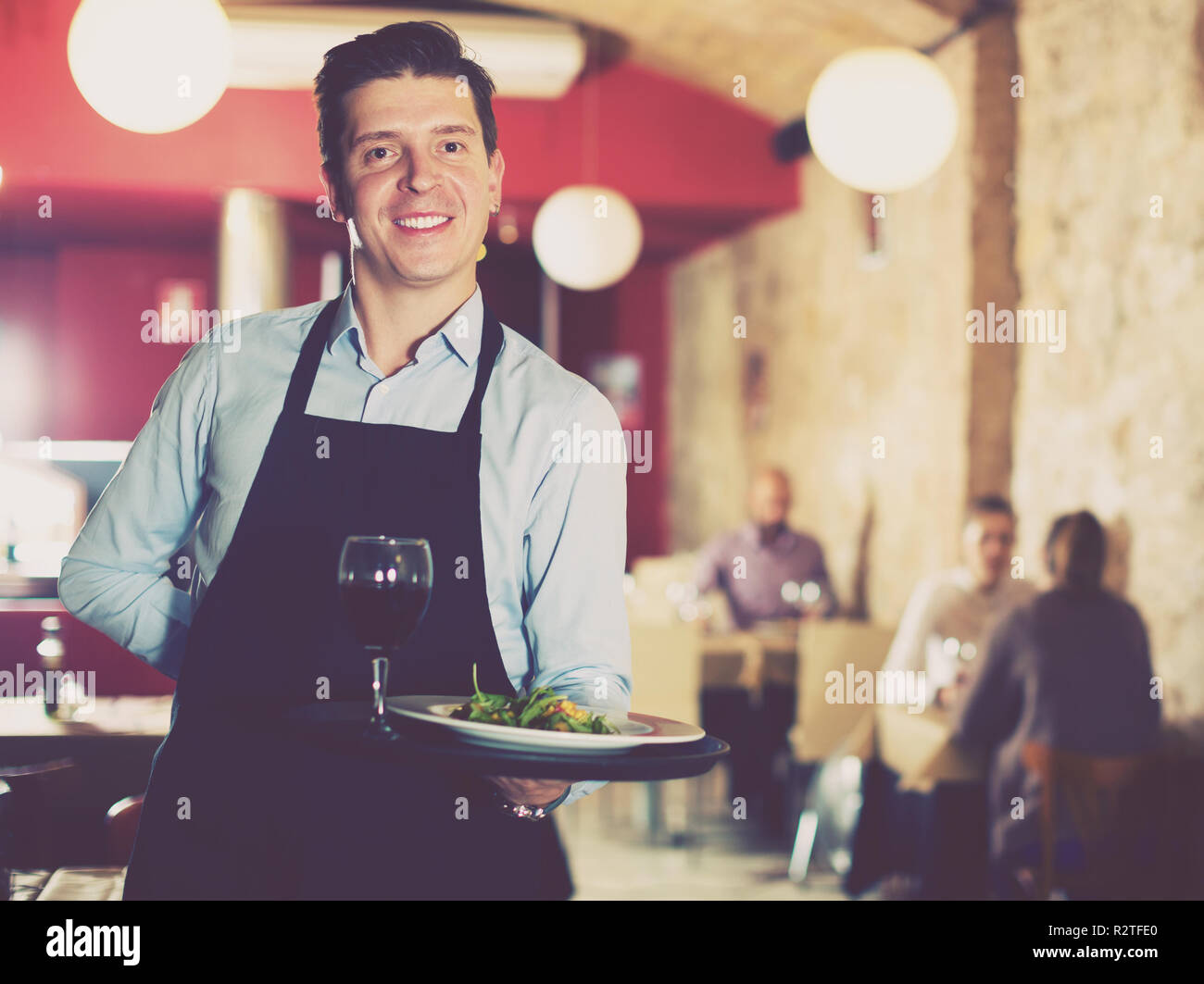 Polite waiter holding tray at restaurant with customers his behind ...