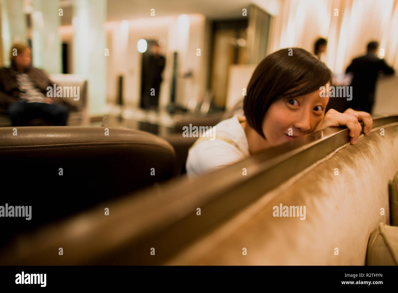 Portrait of a young woman hiding behind a sofa inside a hotel lobby ...