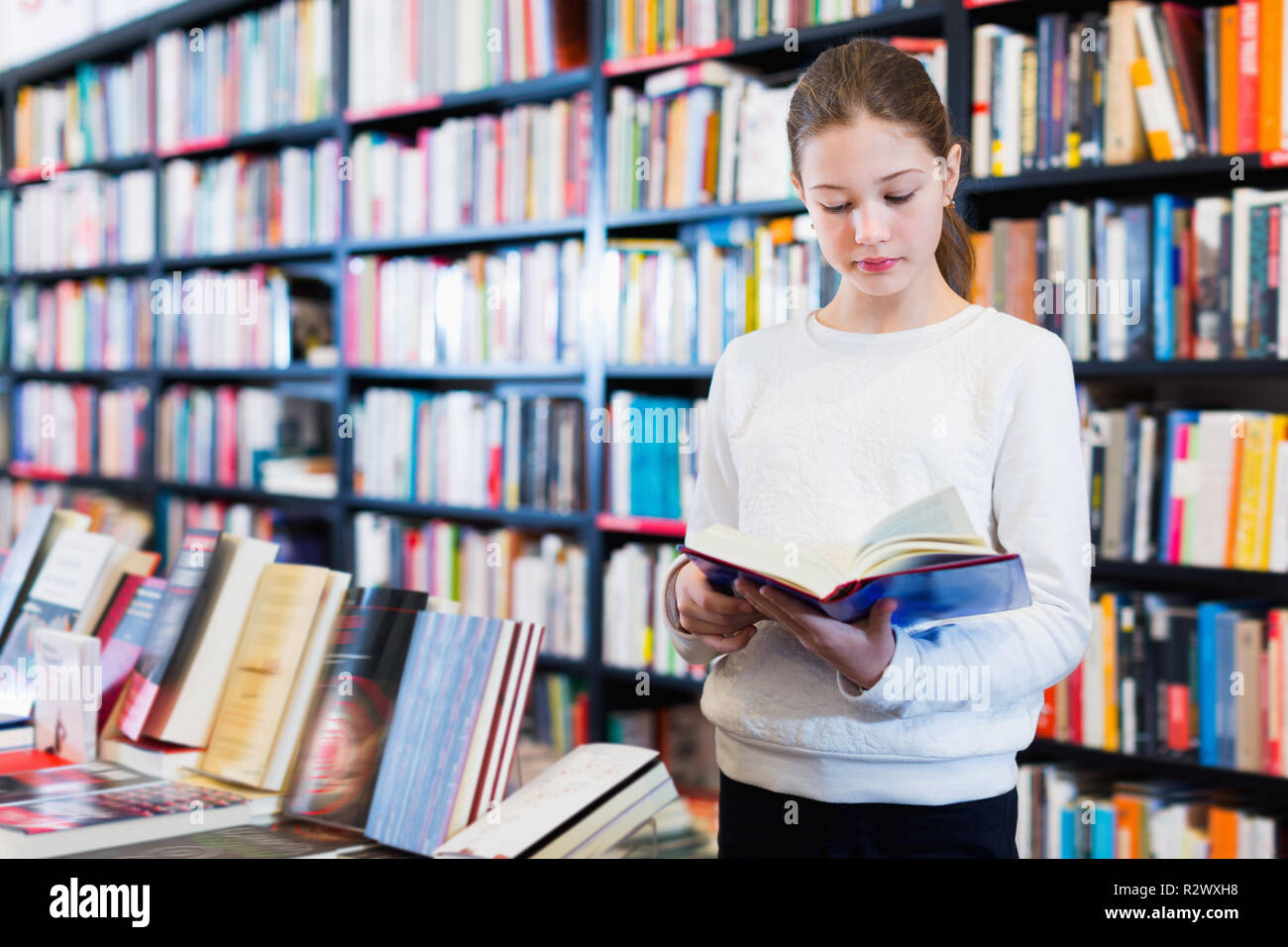 Intelligent positive smiling preteen girl standing alone near bookcase ...