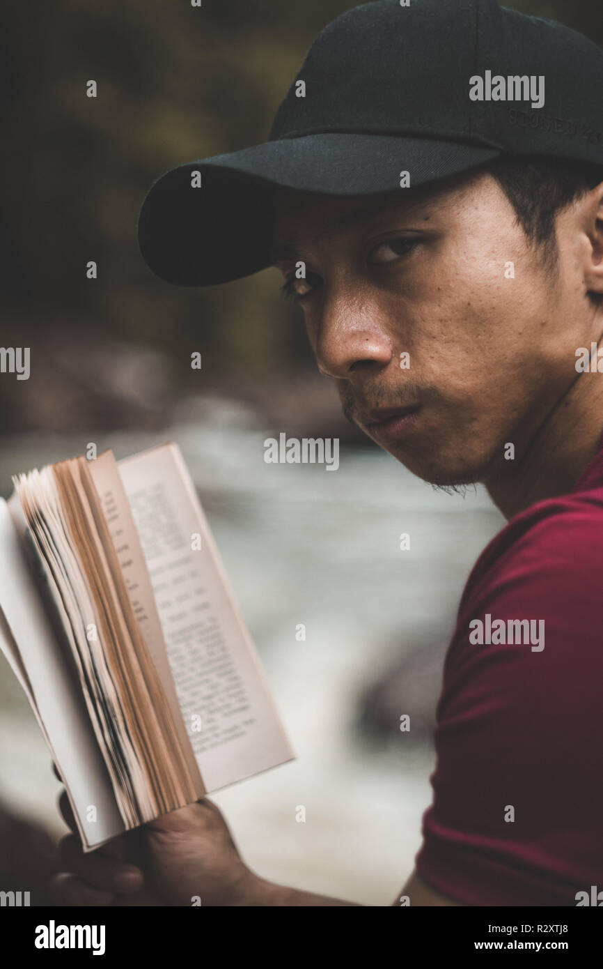 Adventurer guy relaxing and reading a book in the woods next to a river ...