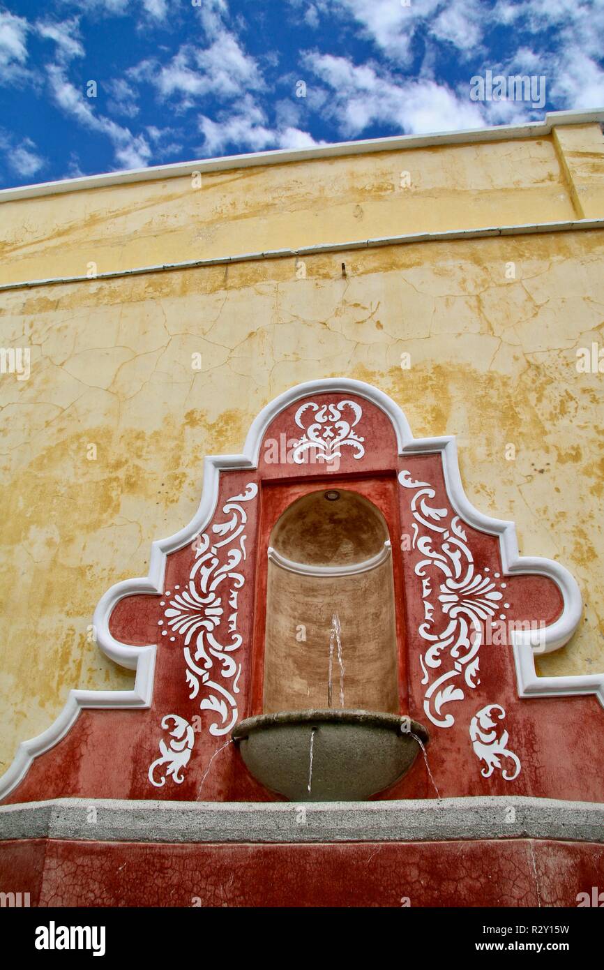 A decorated red fountain on a yellow wall with a blue sky showing ...