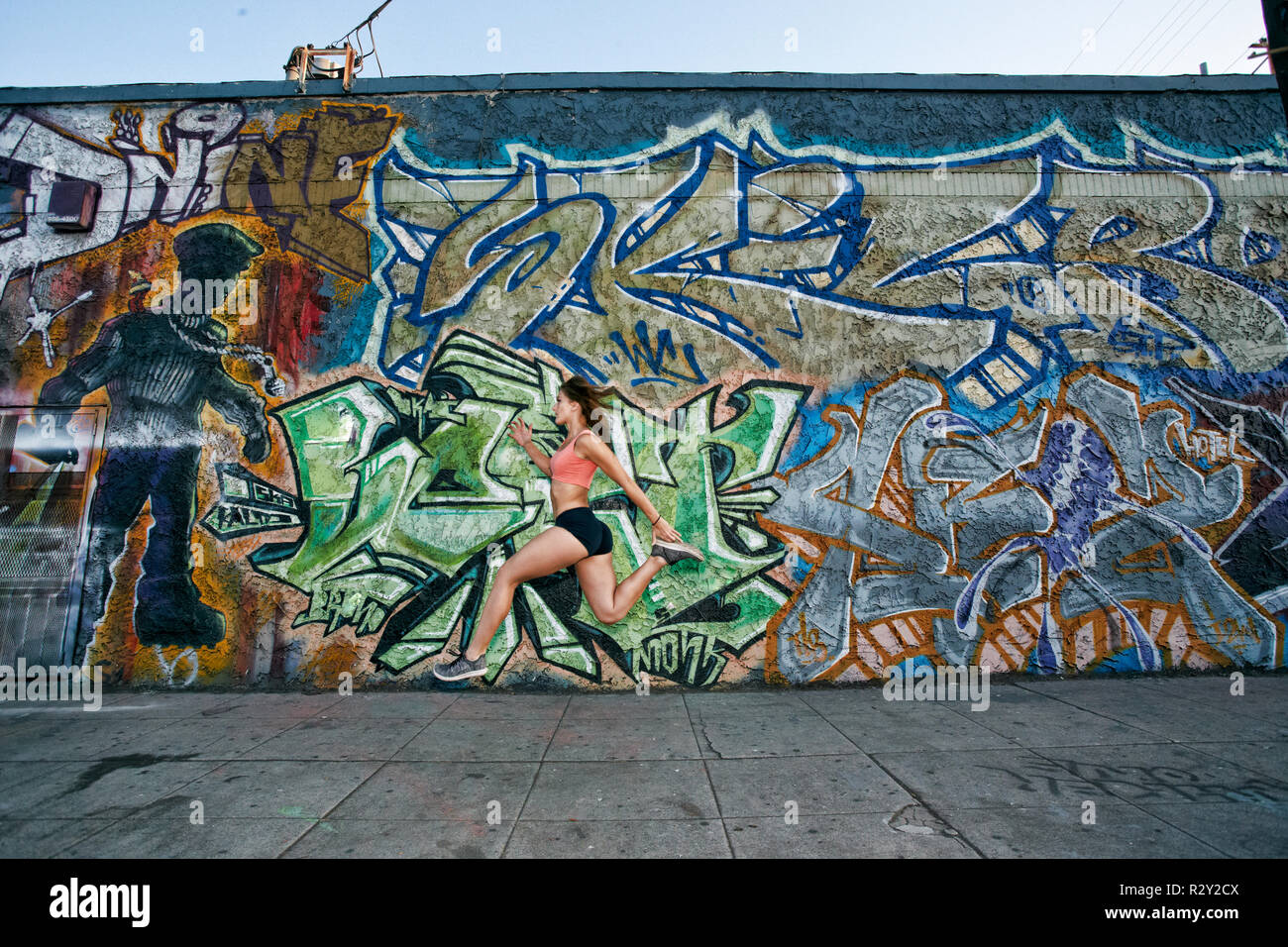 Female athlete running along sidewalk past building covered in graffiti ...