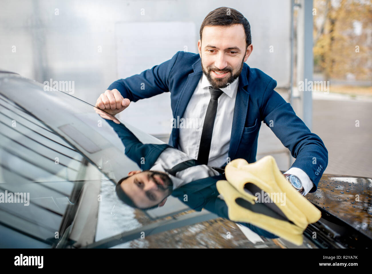Smiling businessman dressed in a suit wiping windshield of his car with ...