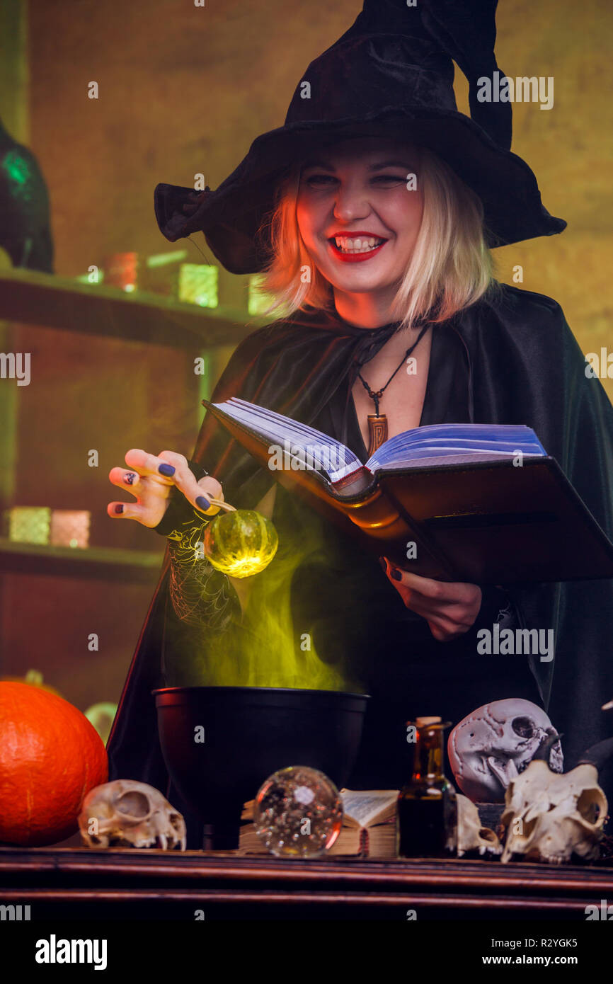 Image of smiling blond witch in black hat with book potting potion in ...