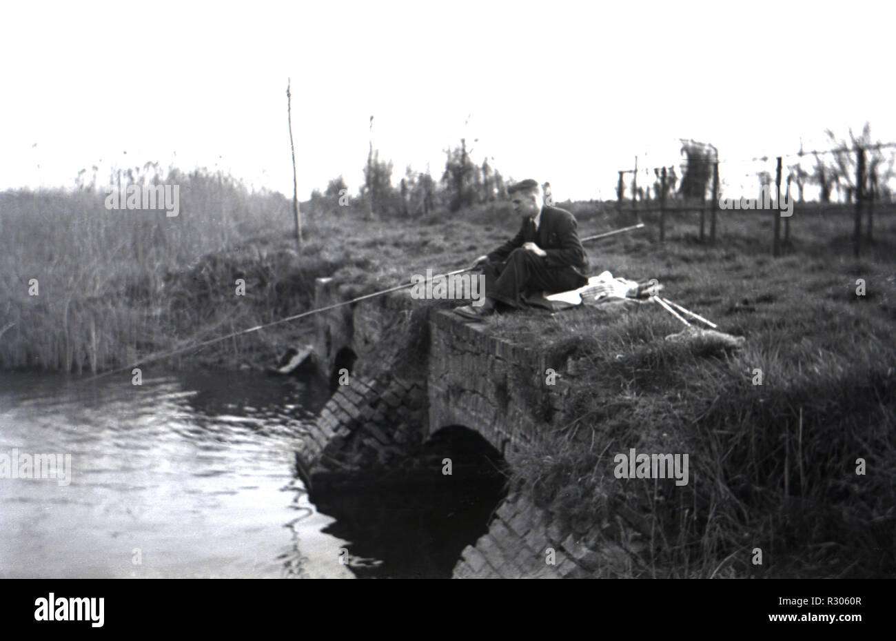 1950s, working man in a suit sitting outside on a riverbank fishing ...