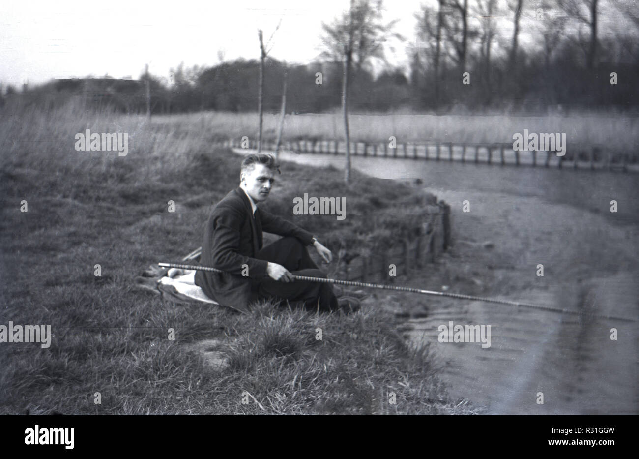 1950s, working man in a suit sitting outside on a riverbank fishing ...