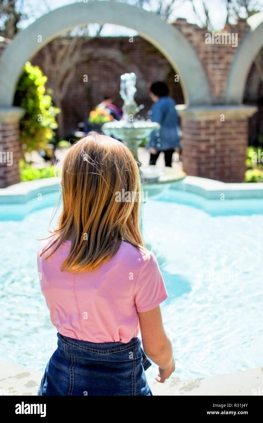 A young blond girl standing by a flowing water fountain and a brick ...