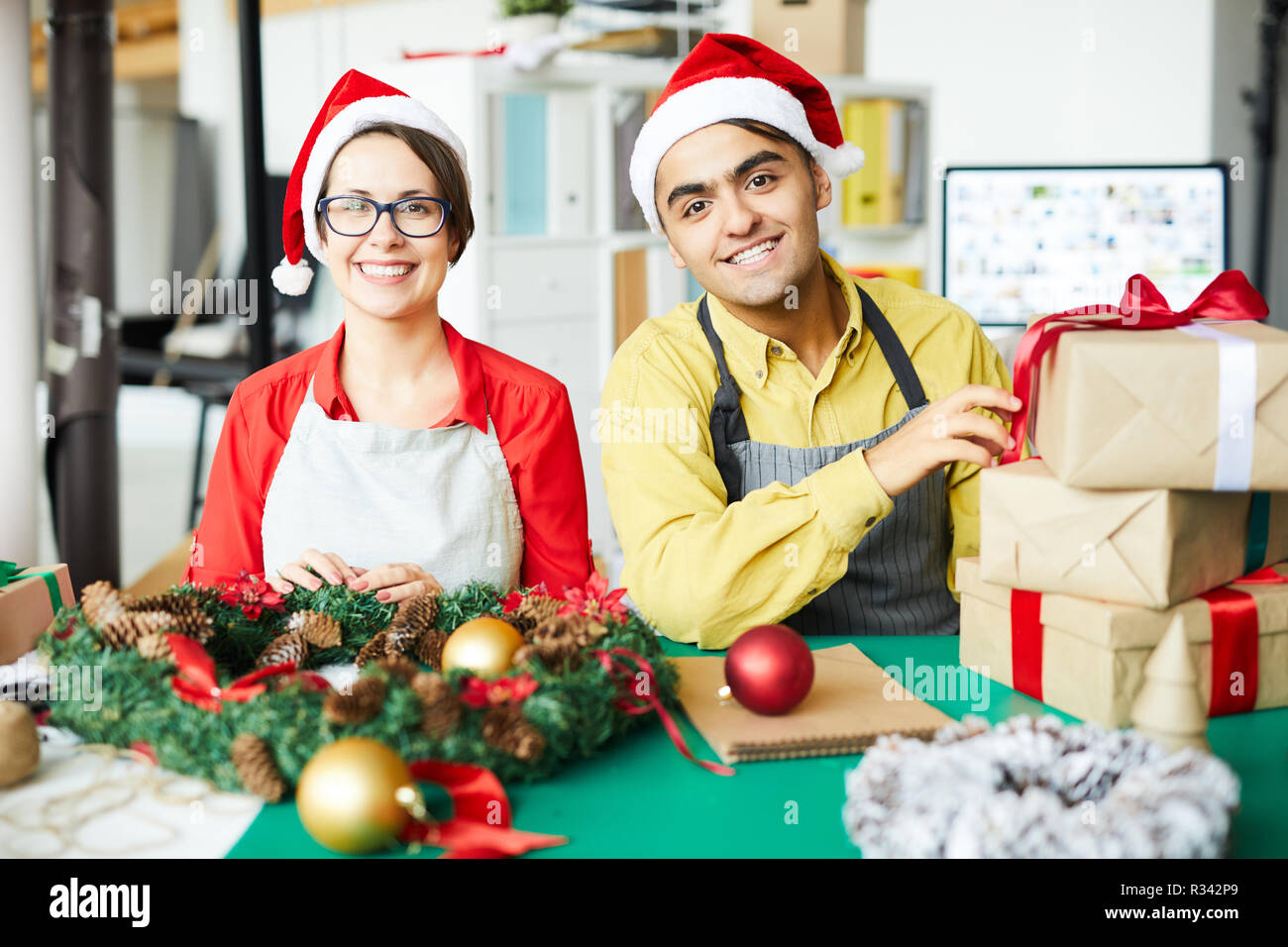 Happy young colleagues in workwear and Santa caps looking at you by ...