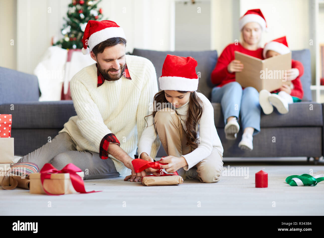 Little Santa girl tying up knot on top of packed giftbox while father ...