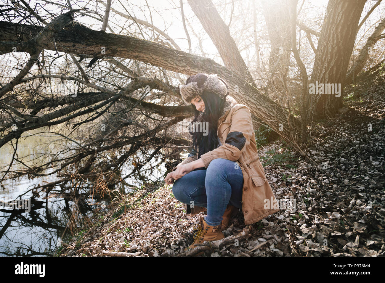 Young brunette woman crouched on the bank of a river wearing a jacket ...