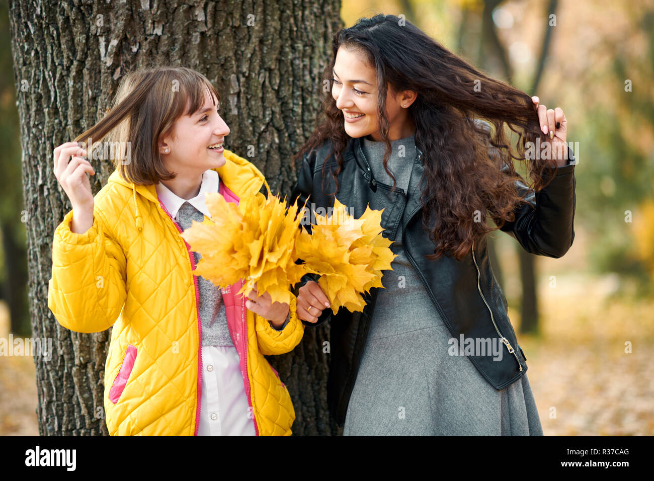 Two girls are in autumn city park. They stand near tree and show hair ...