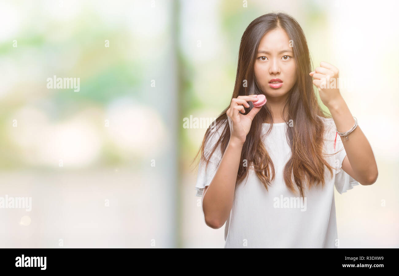 Young asian woman eating pink macaron sweet over isolated background ...