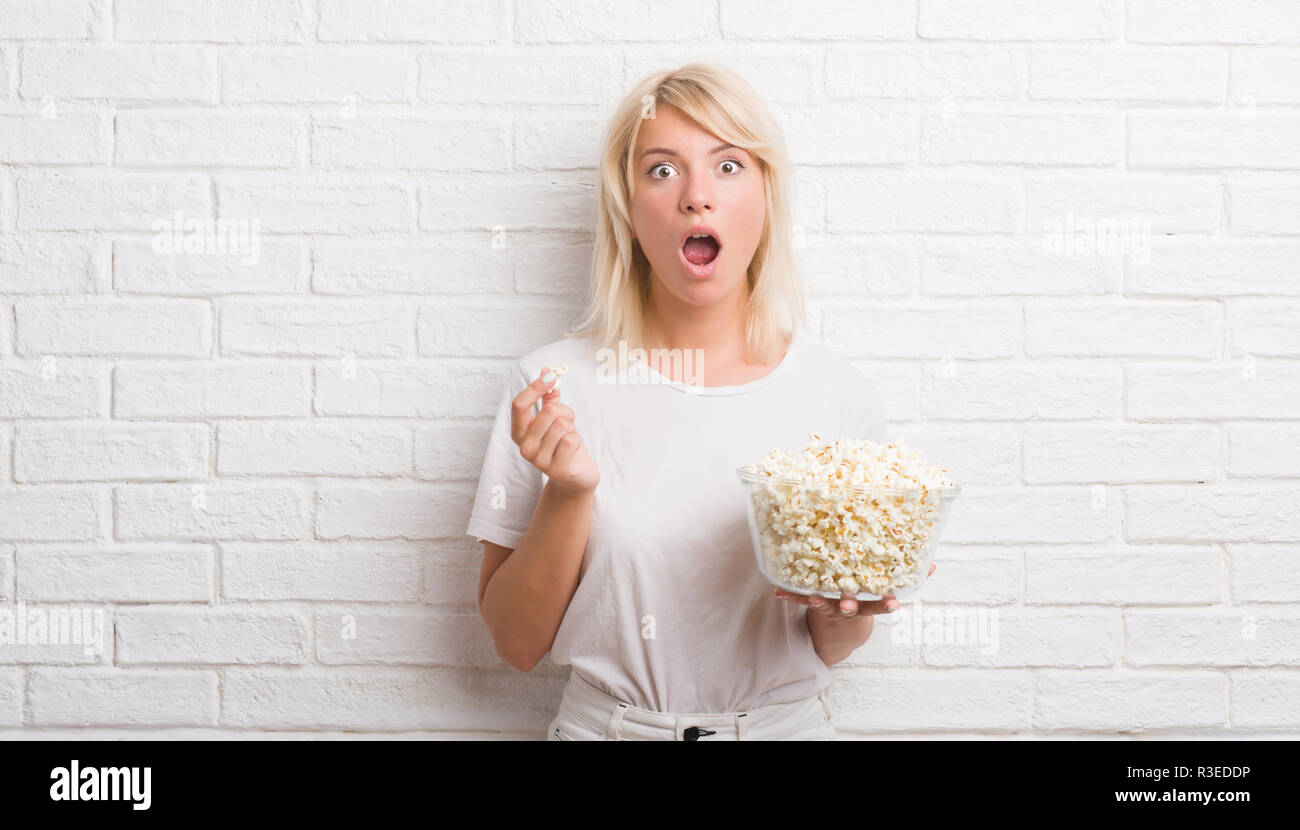 Adult caucasian woman over white brick wall eating pop corn scared in ...