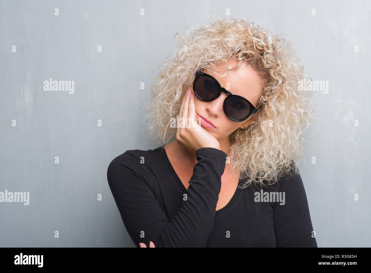 Young blonde woman with curly hair over grunge grey background thinking ...