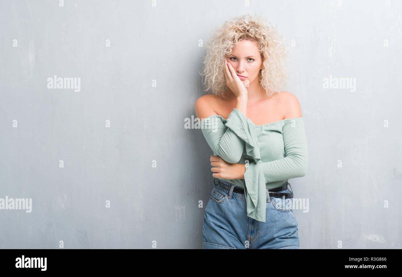 Young blonde woman with curly hair over grunge grey background thinking ...