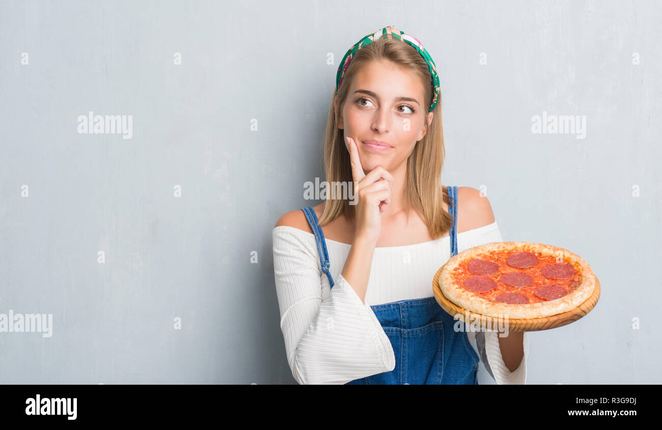 Beautiful young woman over grunge grey wall eating pepperoni pizza ...
