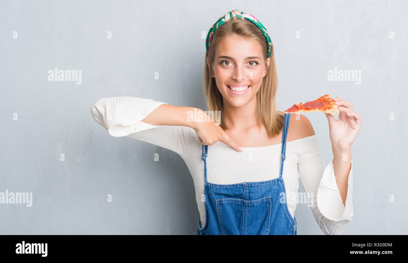 Beautiful young woman over grunge grey wall eating pepperoni pizza ...