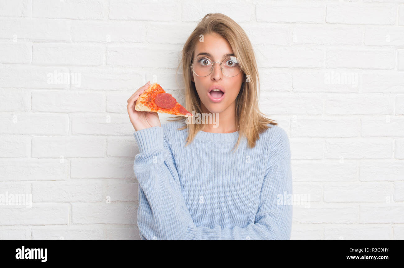 Beautiful young woman over white brick wall eating pizza slice scared ...