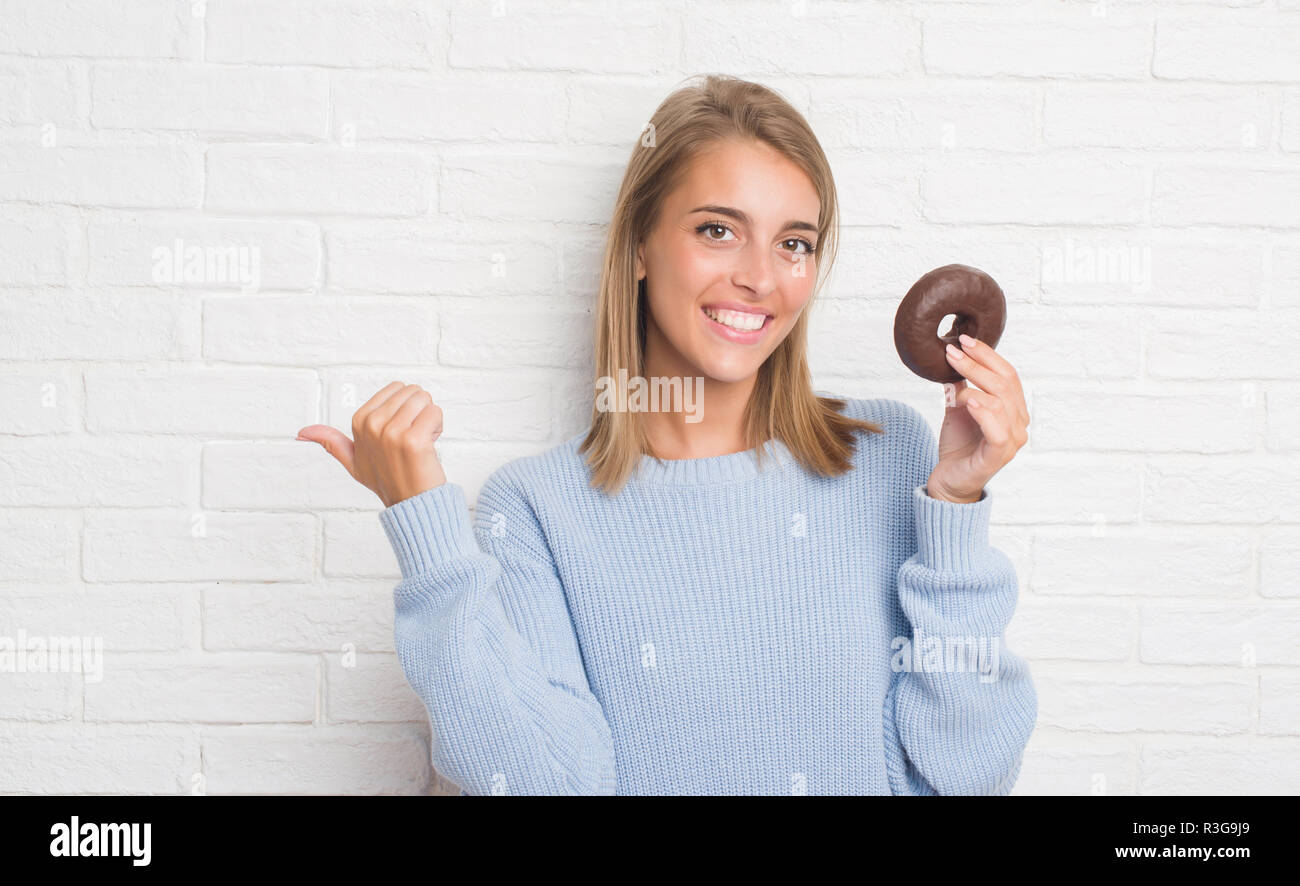 Beautiful young woman over white brick wall eating chocolate donut ...