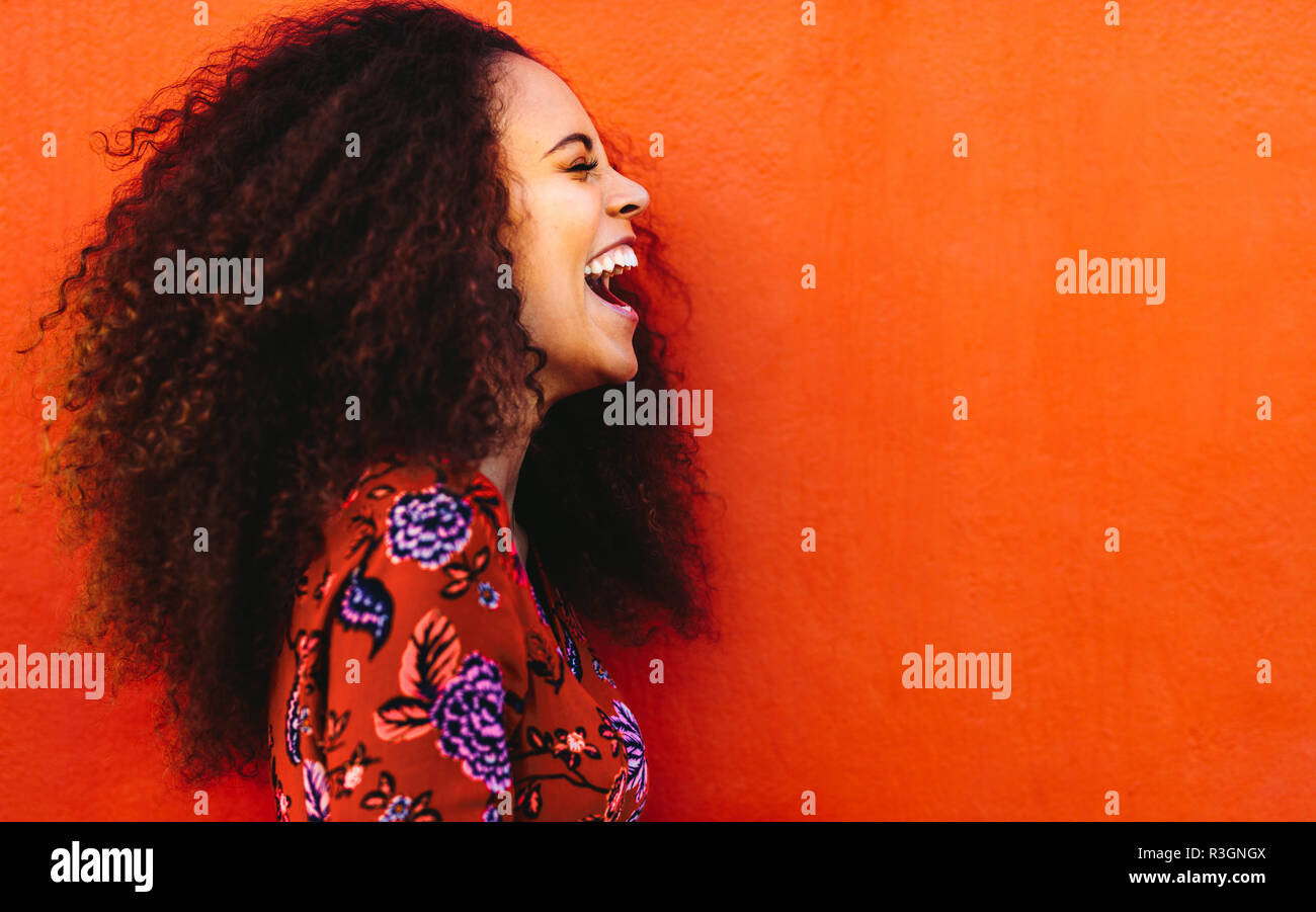 Side view of cheerful woman with curly hair standing against orange ...