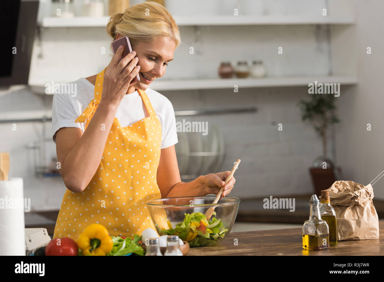 smiling mature woman in apron talking by smartphone while cooking in ...