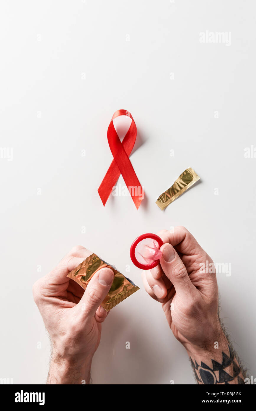 Male Hands Holding Golden Packaging And Red Condom With Aids Awareness Red Ribbon On White