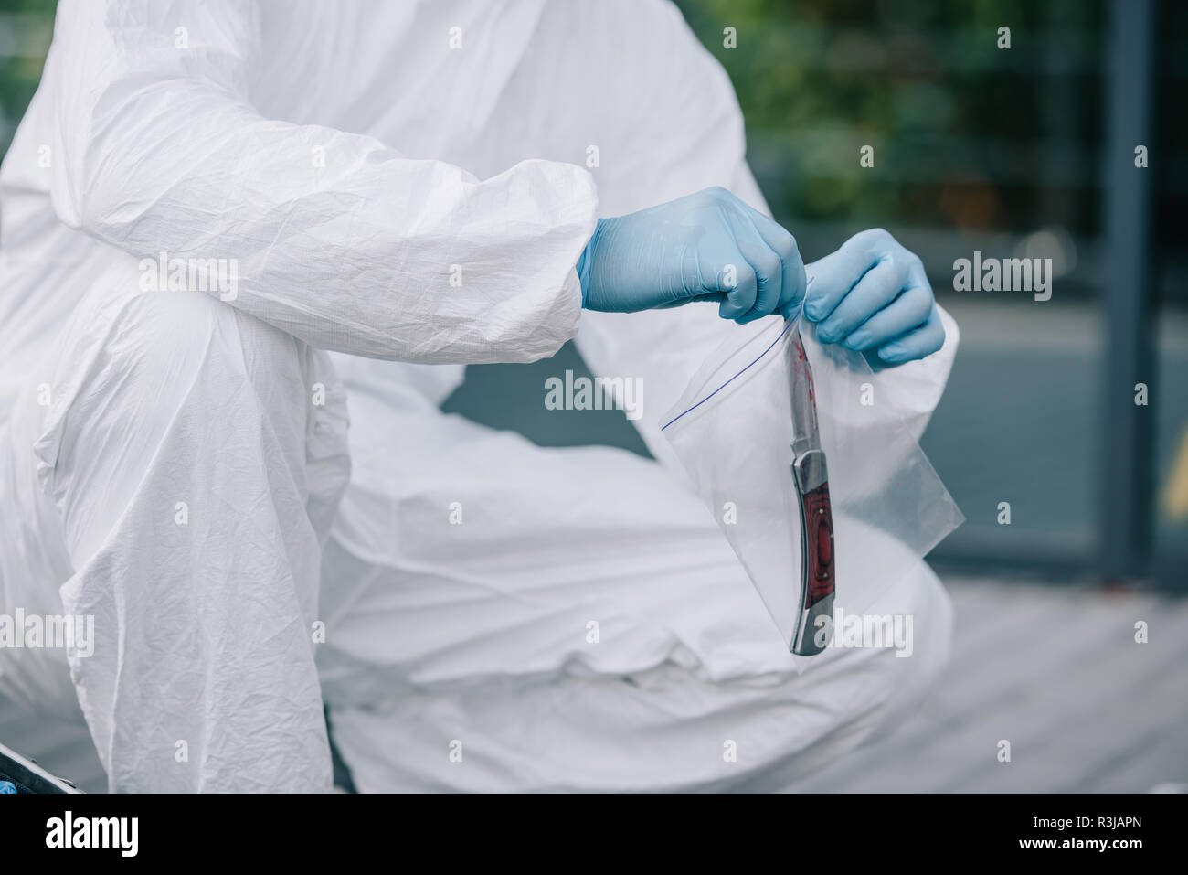 cropped view of criminologist in protective suit and latex gloves taking evidence into flask at ...