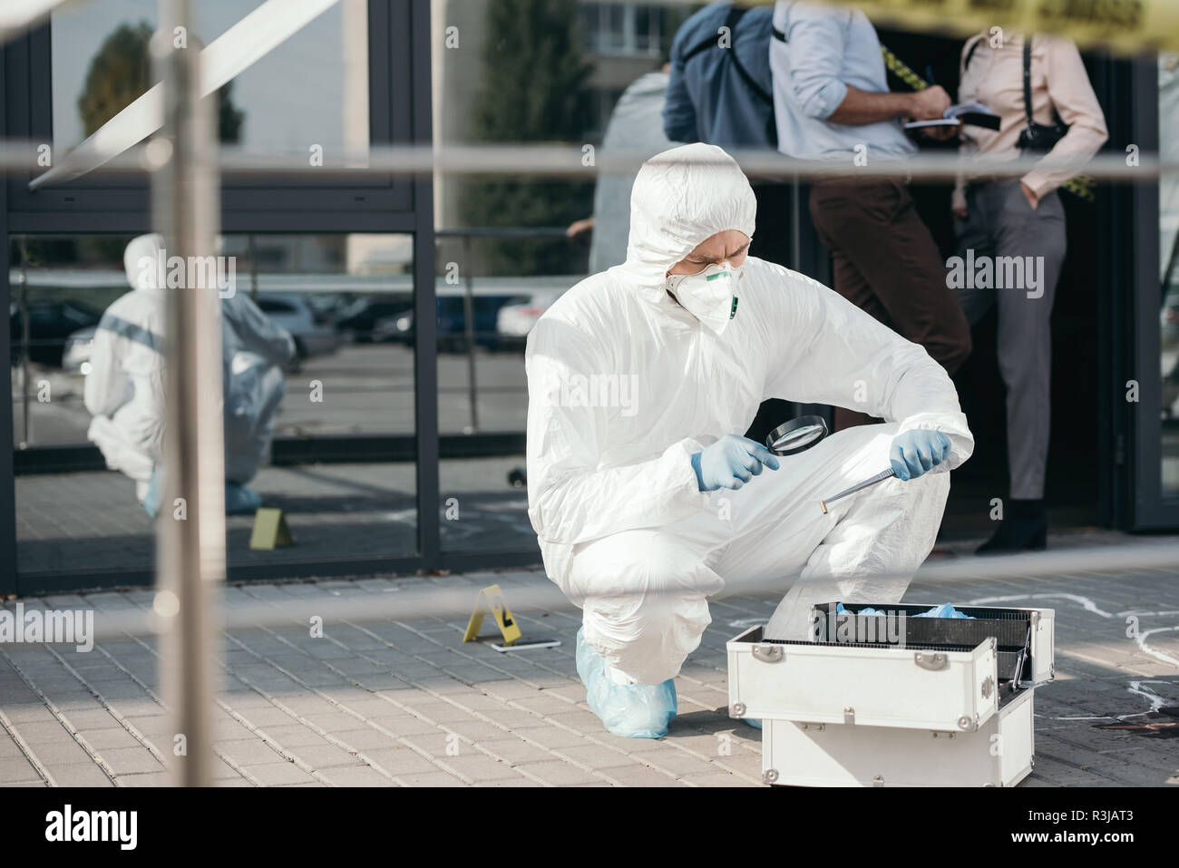 male criminologist in protective suit and latex gloves looking through a magnifier at evidence ...