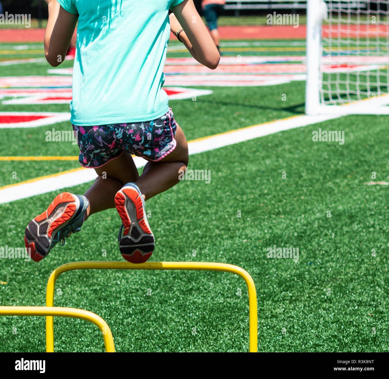 A young girl is jumping over a two foot yellow hurdle on a green turf ...