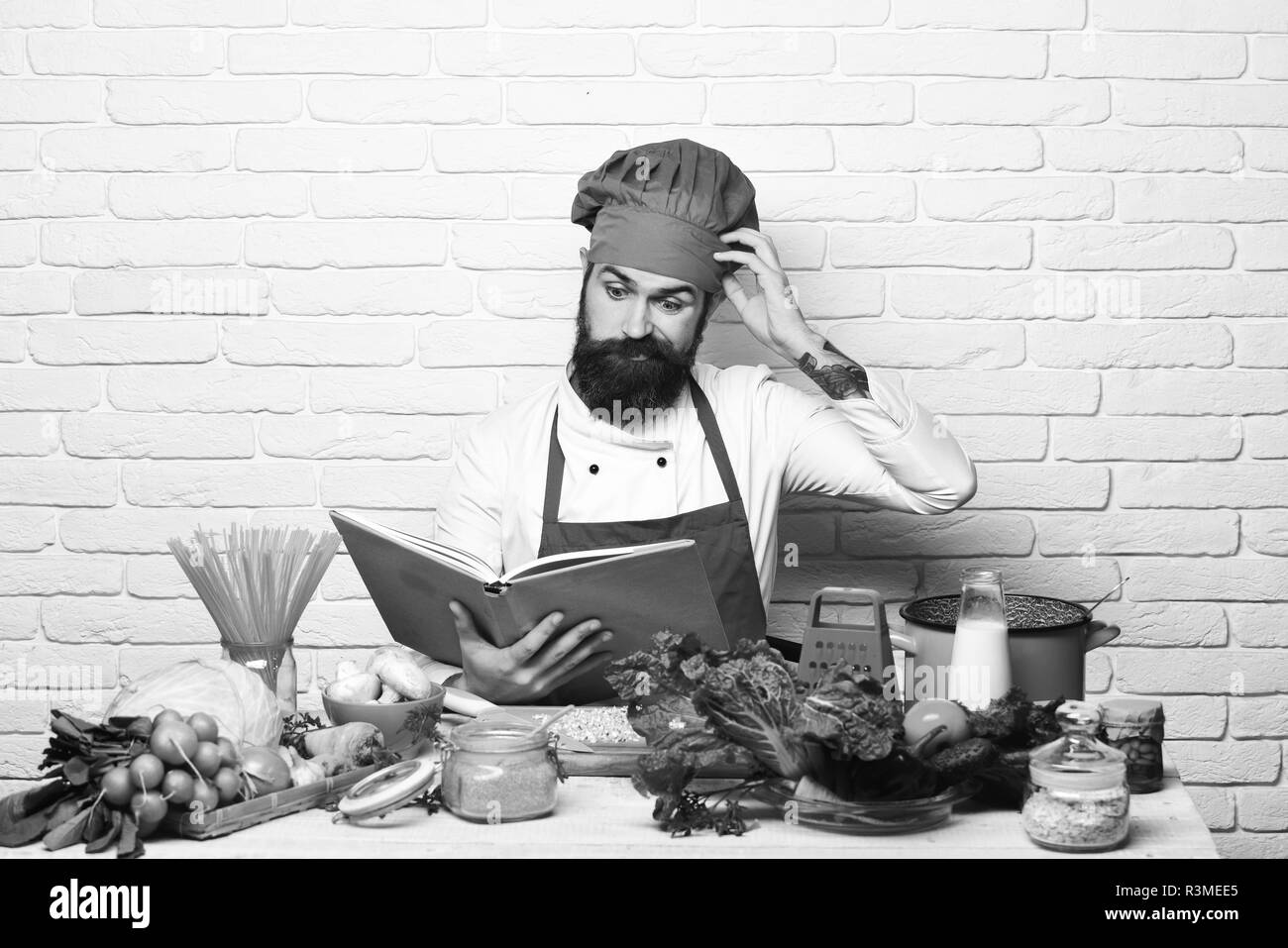 Cook with confused face in uniform sits by table with vegetables, pasta ...