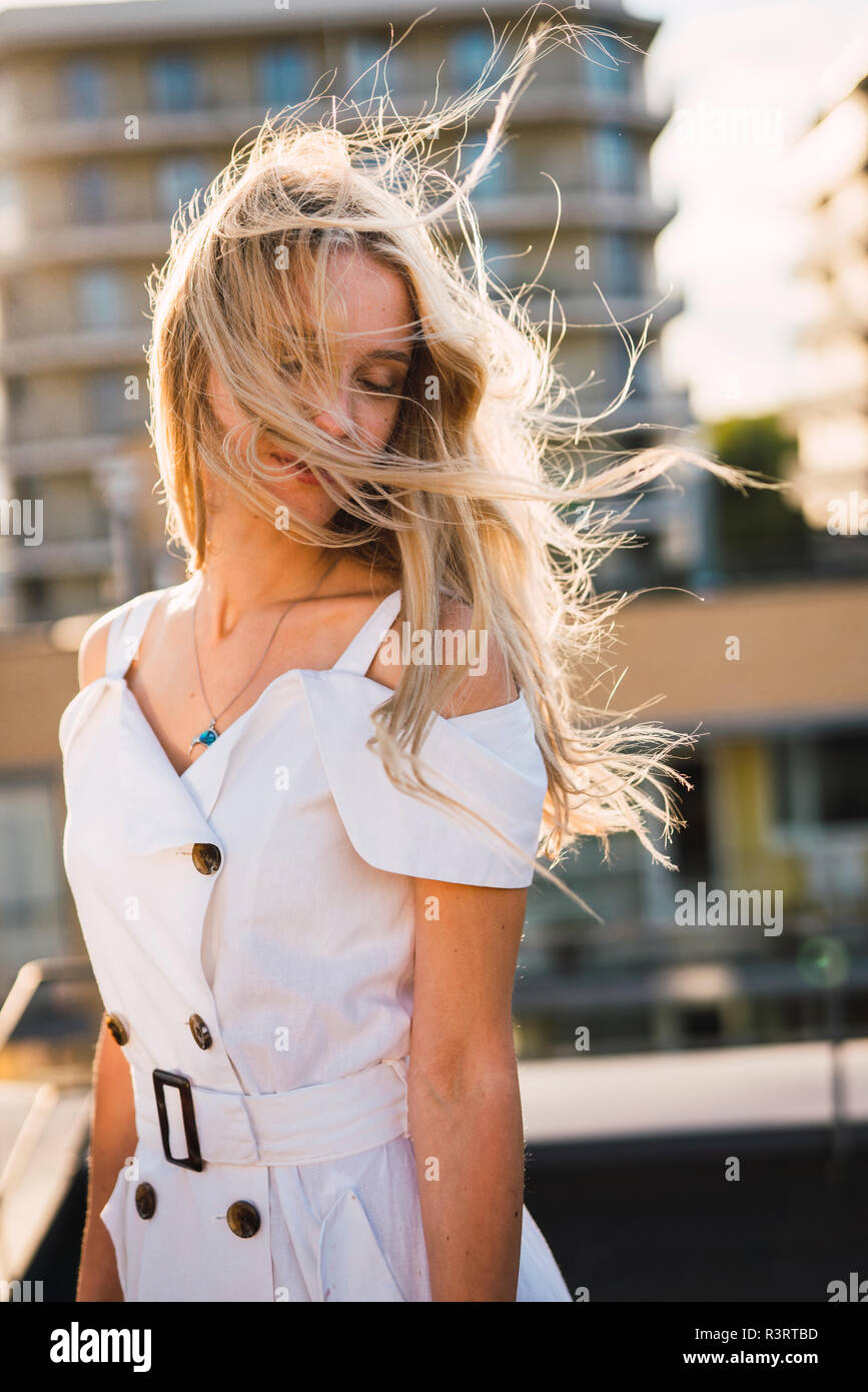 Blond young woman with windswept hair wearing white dress outdoors ...